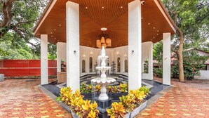 Um pavilhão coberto com uma fonte central, rodeado por plantas em vasos e um chão estampado.