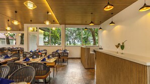 Interior de restaurante com teto de madeira, mesas com cadeiras e grandes janelas com vista para árvores.