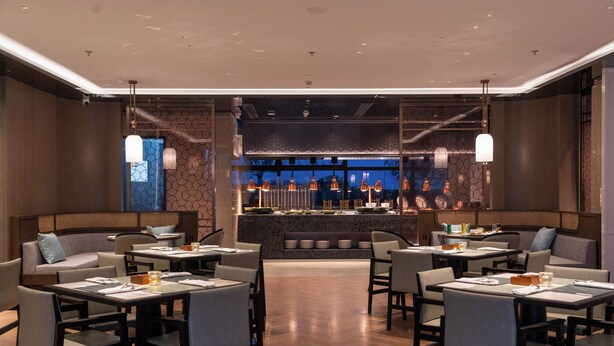 Interior de restaurante com várias mesas e cadeiras, área de bar com garrafas e iluminação pendente.