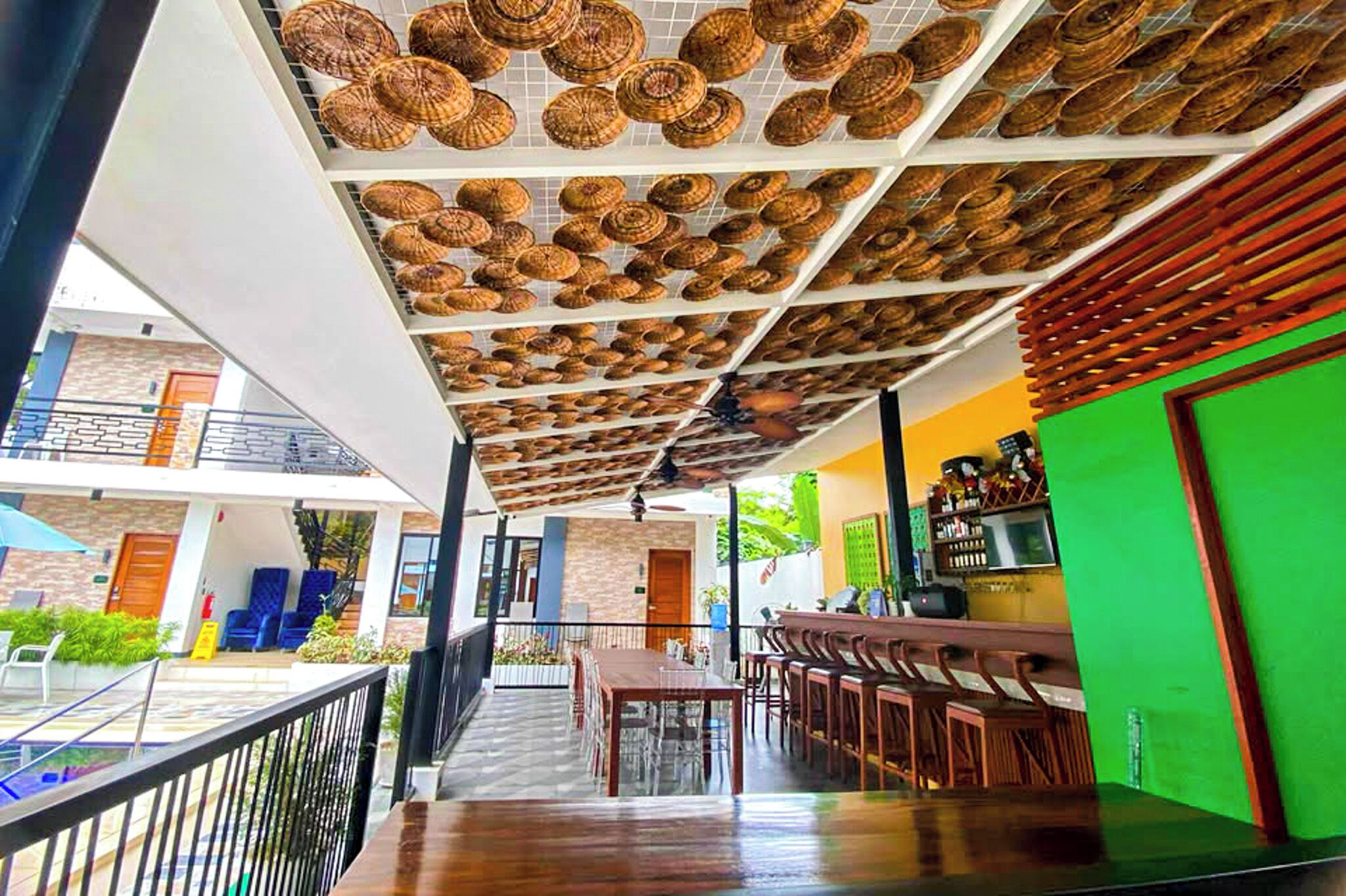 Uma área de estar coberta ao ar livre com piso de madeira, um bar com banquetas e um fundo verde.