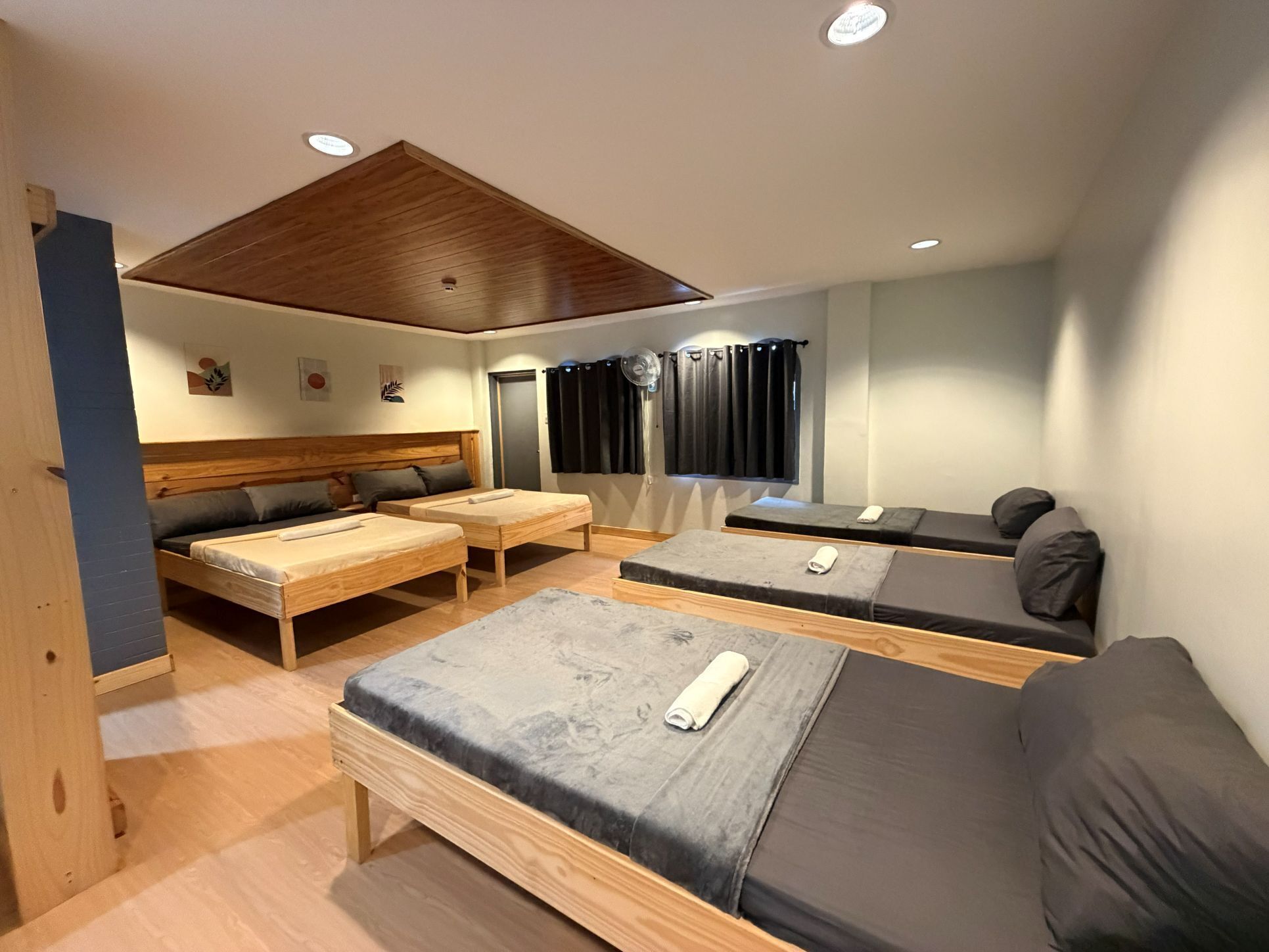 Quarto com duas camas, uma mesinha, um ventilador e janela com cortinas.