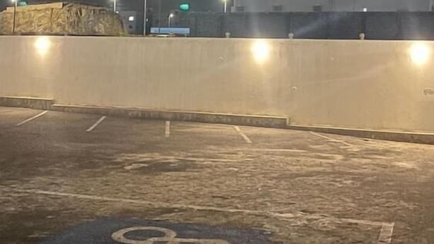 Uma área de estacionamento com o símbolo de estacionamento para deficientes, um edifício ao fundo e luminárias de rua.