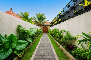 Uma passagem leva a um edifício com telhado de telhas vermelhas, ladeado por vegetação exuberante e uma parede com jardineiras suspensas.