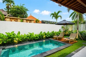 Piscina com parede branca e vegetação exuberante, deck de madeira e área de estar coberta.