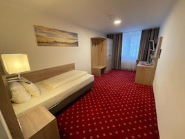 Quarto de hotel com cama, escrivaninha, espelho, tapete vermelho e um quadro na parede.