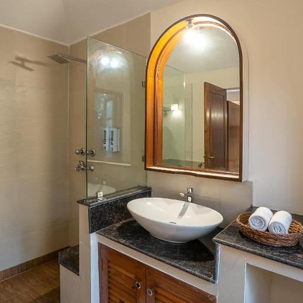Uma casa de banho com cabine de duche em vidro, lavatório branco, espelho com moldura de madeira e cesto com toalhas.