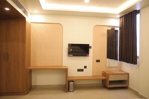 Quarto com televisão na parede, um armário de madeira, uma pequena prateleira e um cesto de lixo.