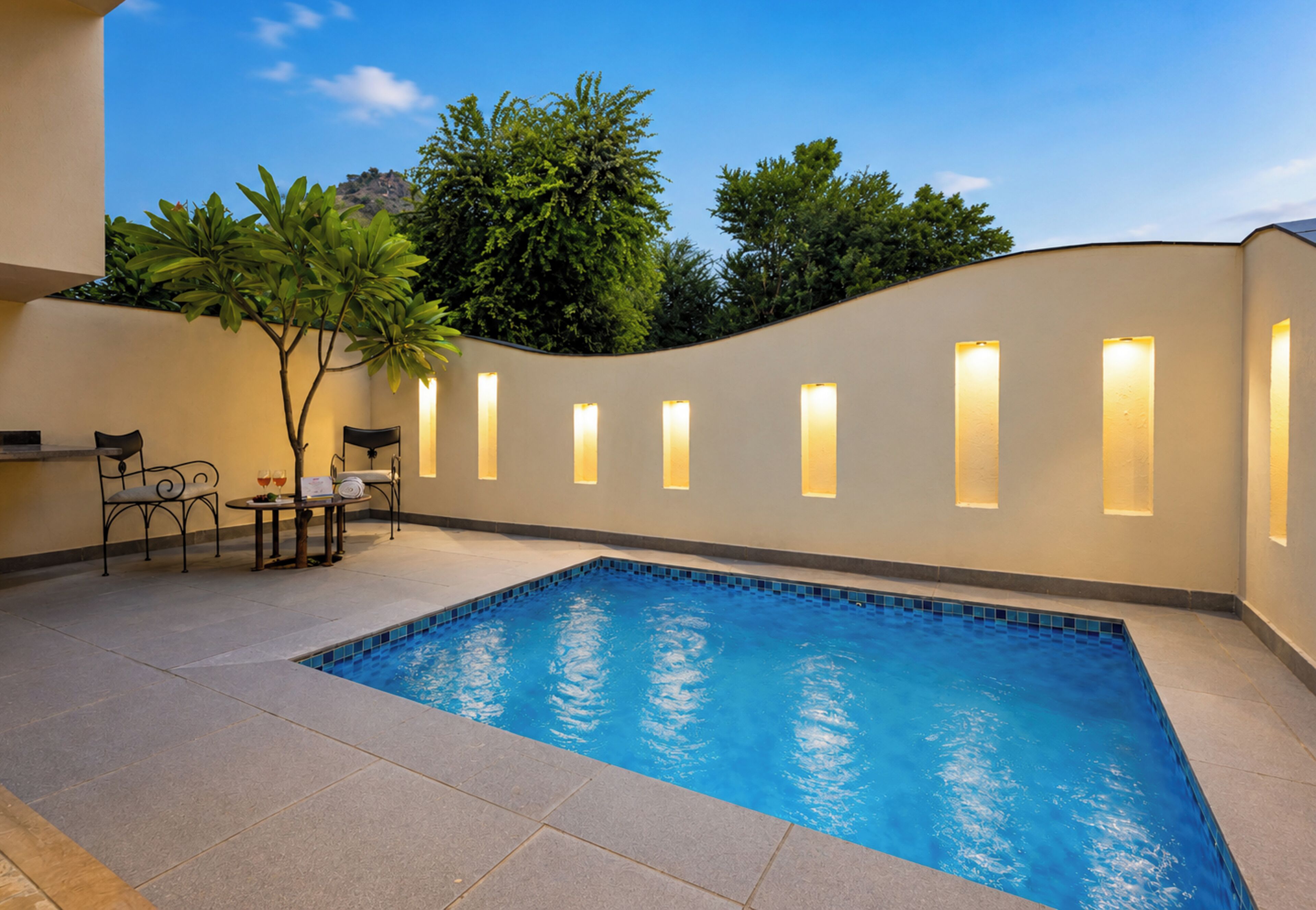 Piscina com deck ladrilhado, área de estar com cadeiras e mesa, e parede com painéis iluminados.