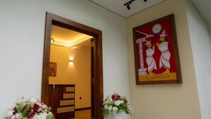 Um corredor com uma porta de madeira que dá para um quarto, uma obra de arte emoldurada na parede e dois vasos com flores.