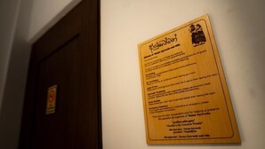 Uma placa de madeira com texto e um logótipo, fixada numa parede num corredor.