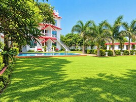 Um edifício grande com piscina, rodeado por relvado bem cuidado e palmeiras.