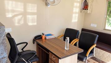 Um pequeno escritório com escrivaninha, duas cadeiras, um ventilador e uma estante com objetos decorativos.