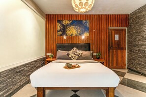 Quarto de hotel com cama, mesa de cabeceira, luminária na parede, porta e uma grande pintura abstrata.