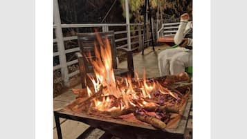 Uma lareira com lenha em brasa, rodeada por cadeiras e uma mesa, localizada em área externa com grades e árvores ao fundo.