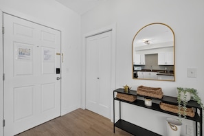 Um corredor com portas brancas, uma mesa consola preta com cestos, um espelho e uma cozinha visível através de uma porta aberta.