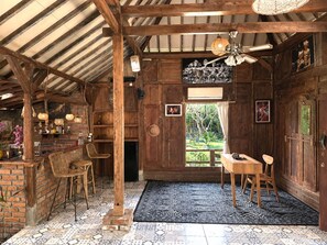 Interior rústico com vigas de madeira aparente, parede de tijolos, mesa de madeira, área de bar e vista para o verde lá fora.