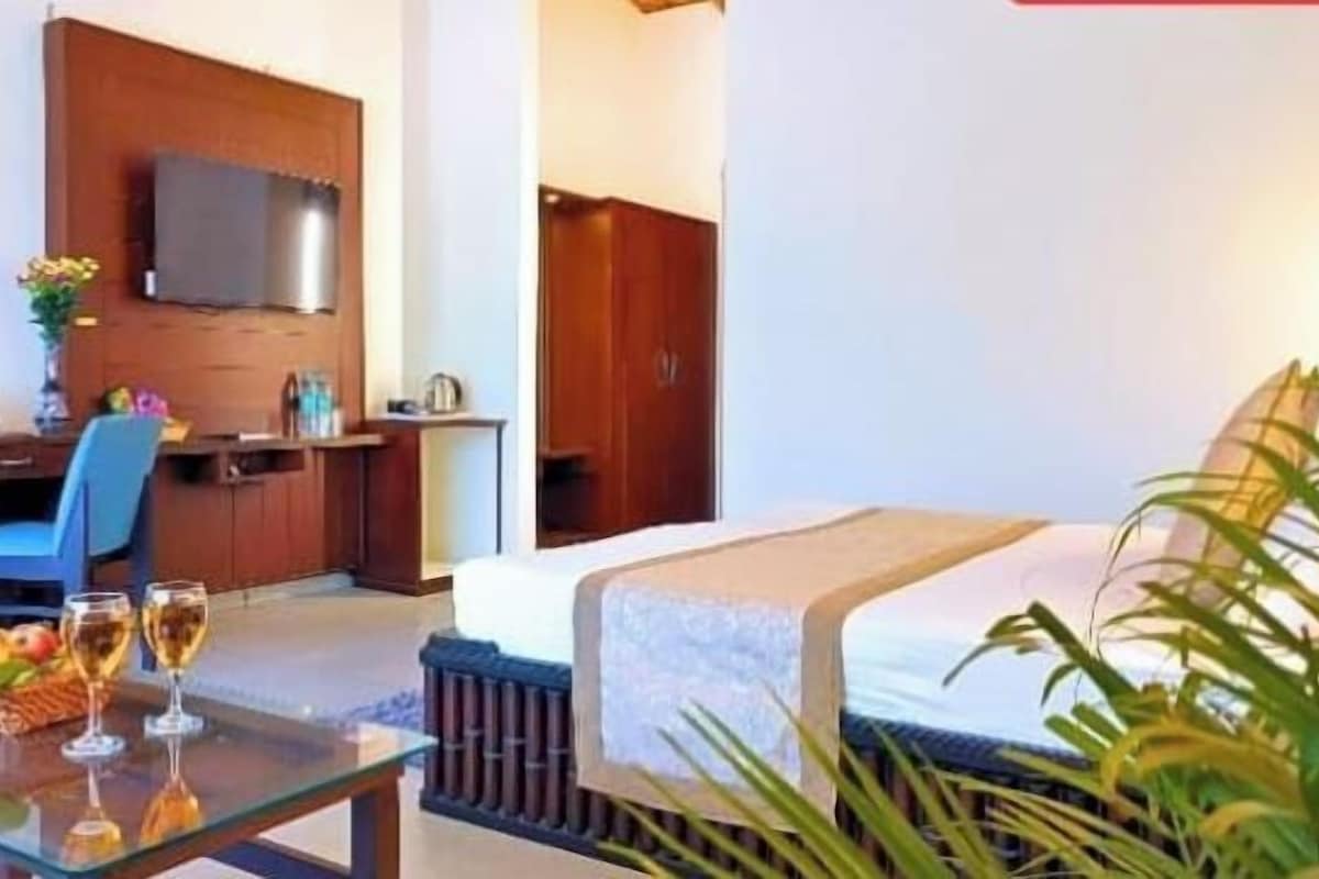 Quarto de hotel com cama, escrivaninha com laptop, cadeira, televisão fixada na parede e mesinha com fruteira e copos.