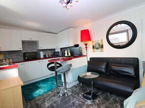 Um espaço de estar compacto com cozinha compacta, um sofá de couro preto, uma mesa redonda e um abajur vermelho.