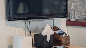 Televisão de tela plana presa na parede, um dispositivo branco sobre uma escrivaninha de madeira, um controle remoto e uma caixa de madeira com etiqueta.