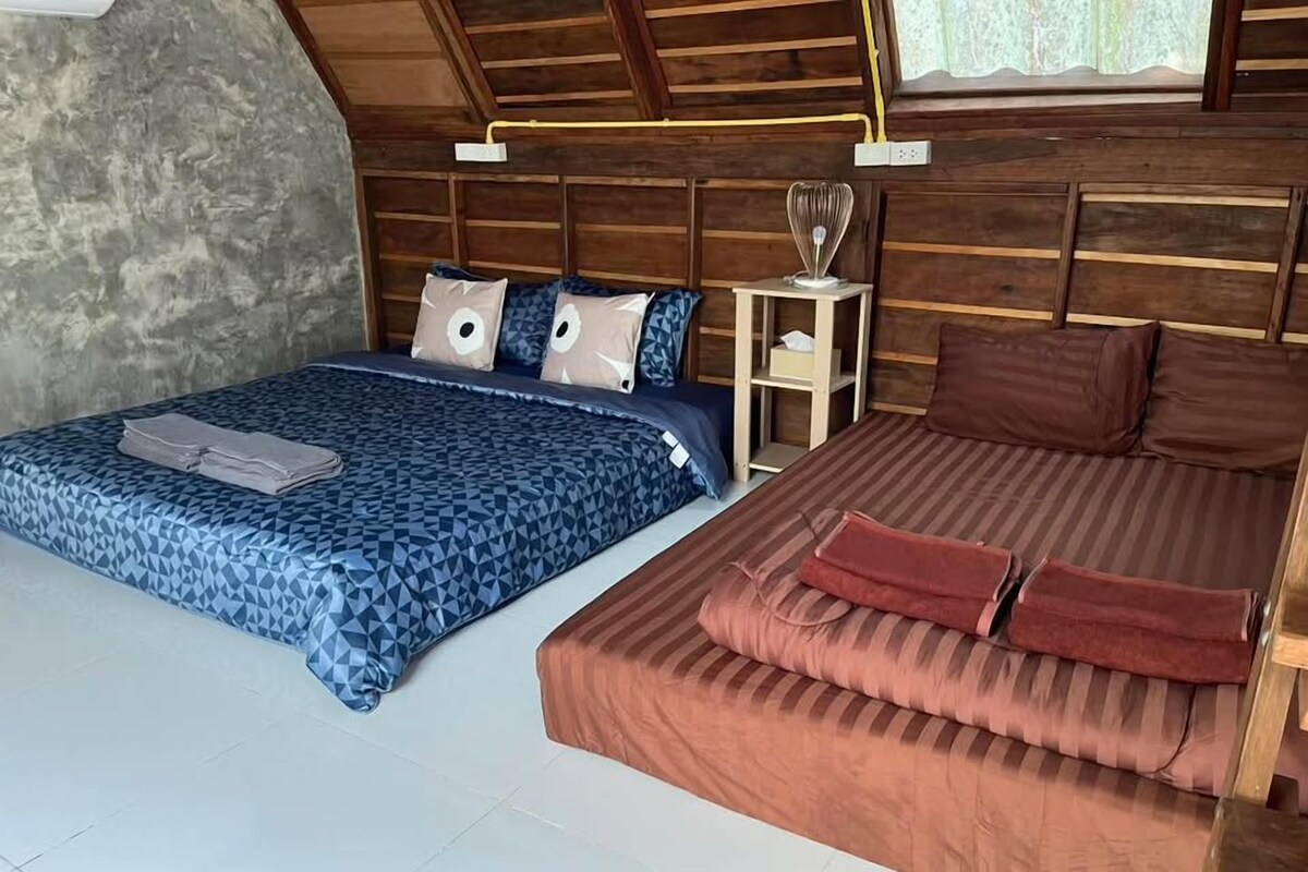 Quarto com duas camas, uma com colcha estampada e outra com colcha listrada. Há uma mesa pequena com abajur e uma prateleira com um objeto decorativo.