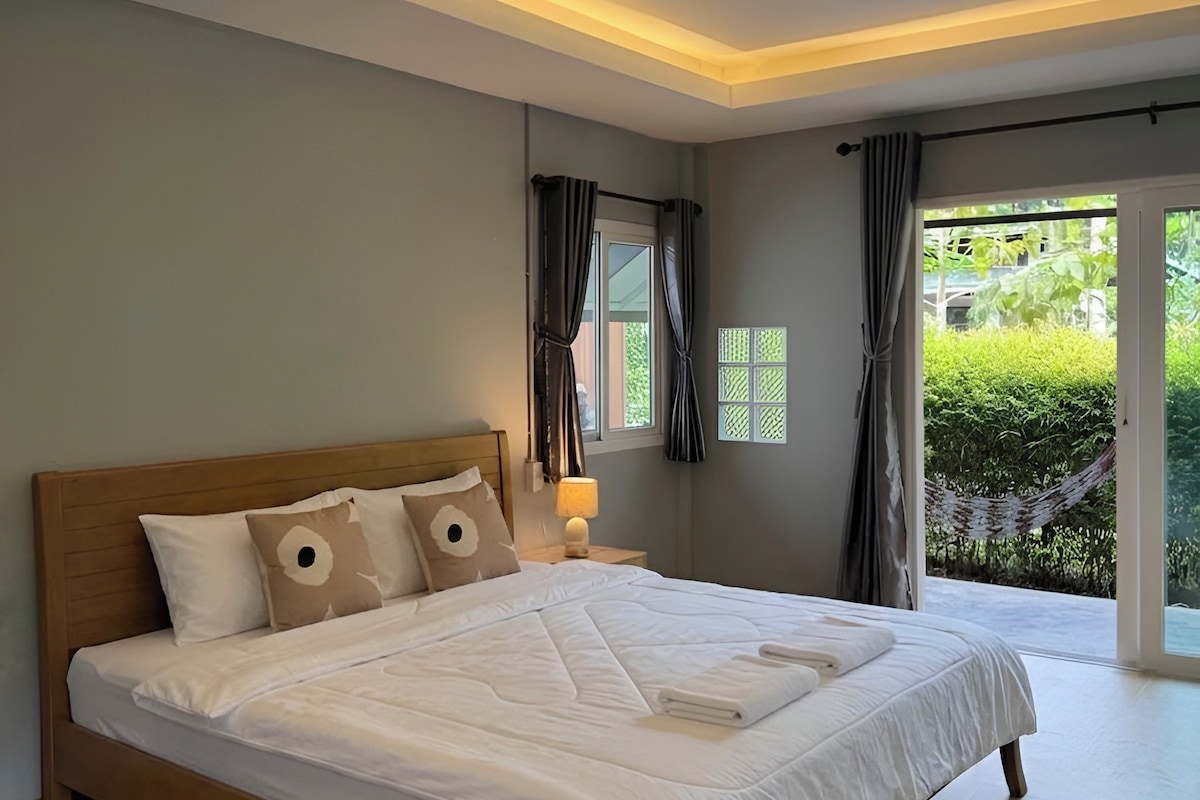 Quarto espaçoso com cama grande, cabeceira de madeira e vista para o verde pela janela.