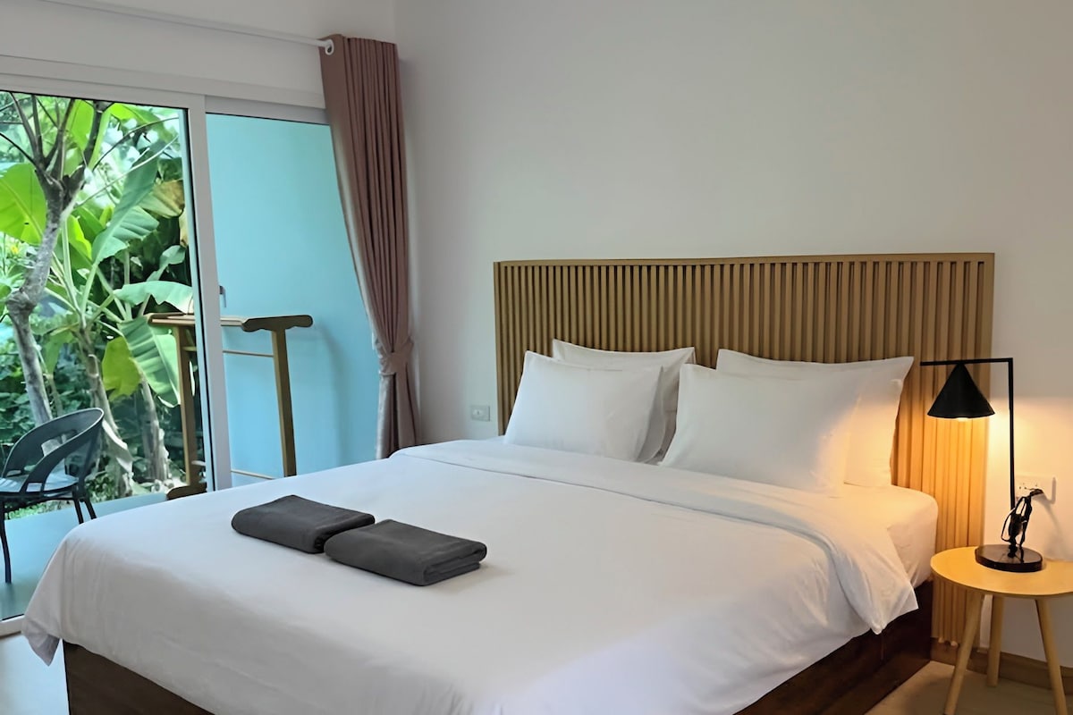 Quarto de hotel moderno com uma cama grande, mesa de cabeceira, luminária e vista para o verde lá fora.