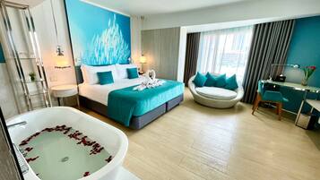 Quarto de hotel moderno com cama grande, área de estar separada, banheira com pétalas de rosa e vista para a cidade.