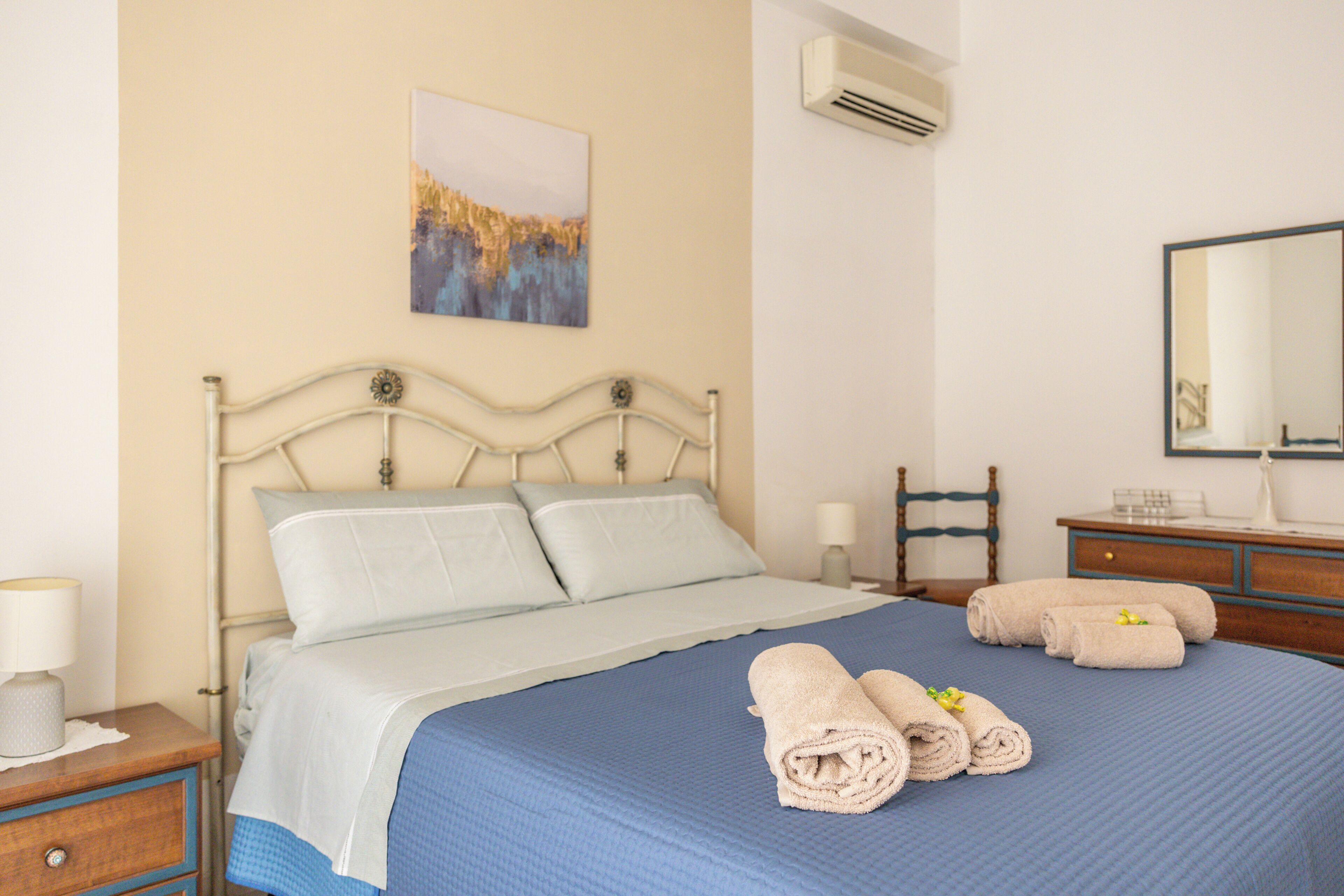 Cama bem arrumada com colcha azul e duas toalhas enroladas, mesa de cabeceira com abajur, uma cadeira e um quadro na parede.
