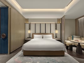 Quarto de hotel moderno com uma cama grande, mesas de cabeceira, área de estar e um painel decorativo na parede.