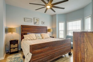Quarto com cama de madeira, duas luminárias de cabeceira, um ventilador de teto e vista para o exterior pelas janelas.