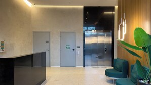 Uma área de recepção com balcão, duas portas, um elevador moderno e um espaço de estar com um vaso de planta.