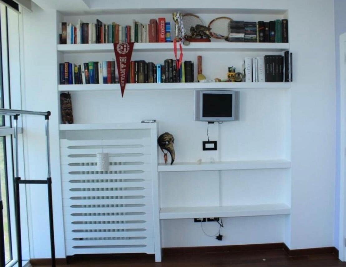 Um quarto com uma estante branca, uma televisão fixada na parede e uma janela com persianas.