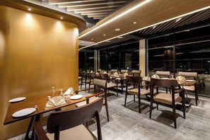 Um restaurante com mesas preparadas para refeições, com decoração moderna e detalhes em madeira, e vista para a paisagem urbana.