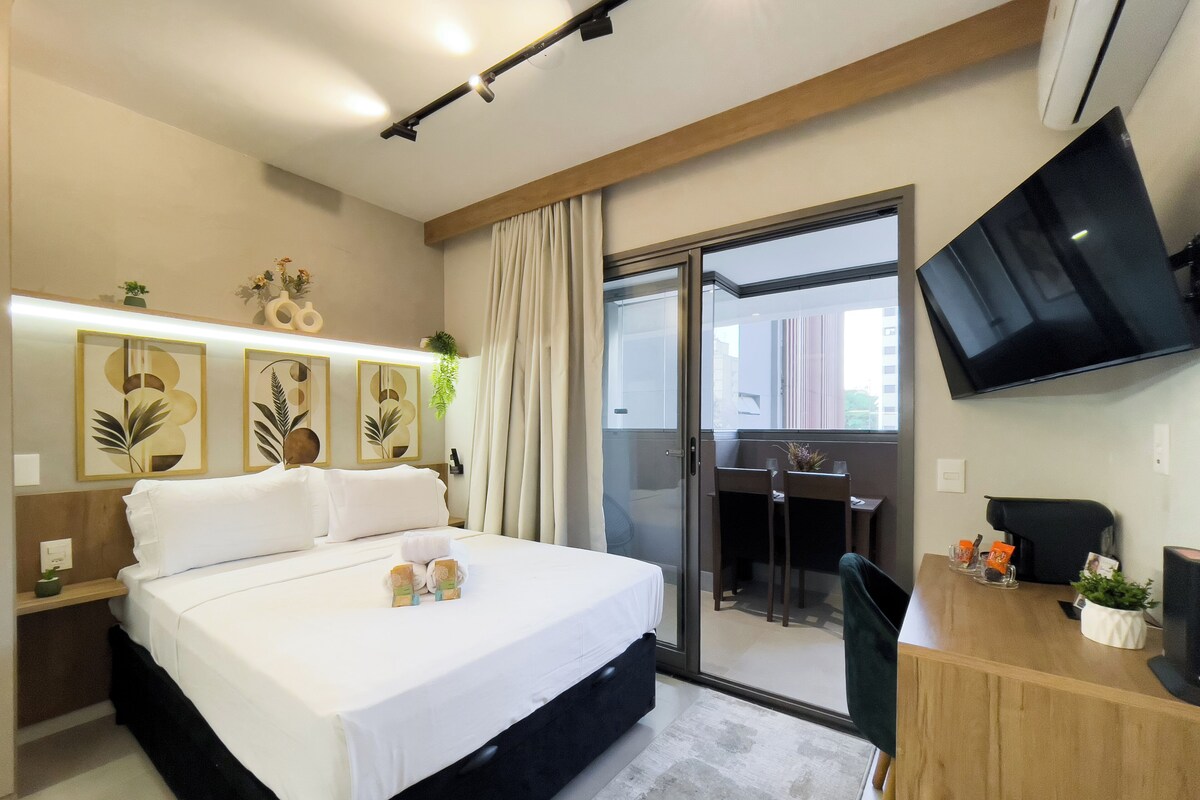 Quarto de hotel com cama, escrivaninha, televisão e porta de correr para uma varanda.