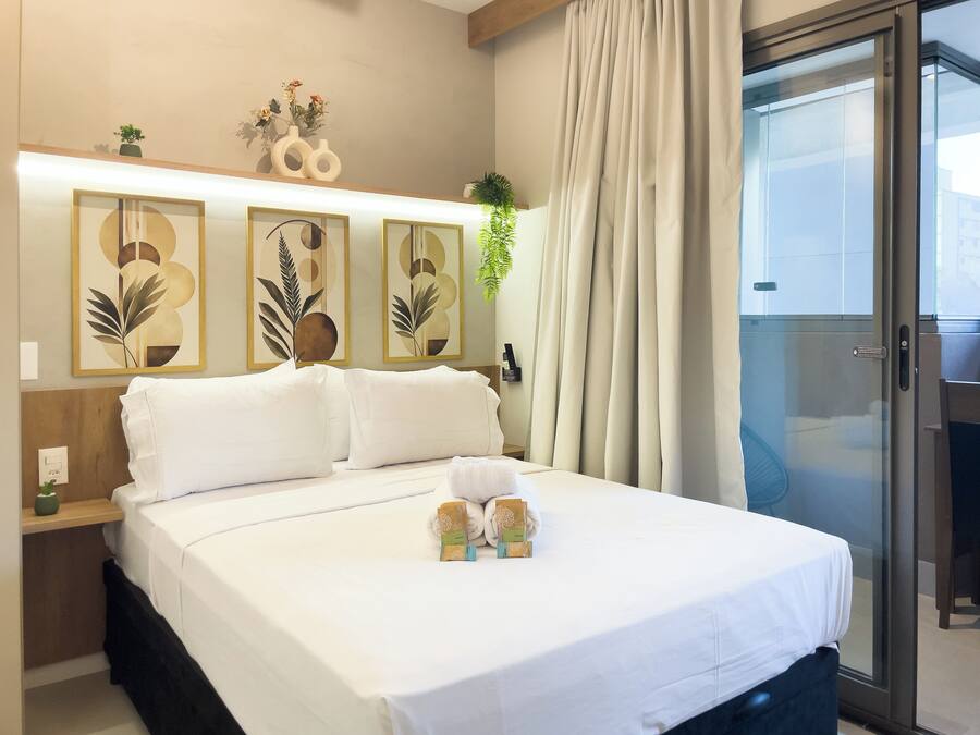 Quarto de hotel com cama, mesa de cabeceira, relógio fixado na parede e janela com cortinas.
