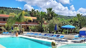 A resort pool with multiple lounge chairs and umbrellas, surrounded by lush greenery and a mountainous backdrop.