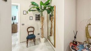Um corredor com uma cadeira de madeira, uma grande planta em vaso, um espelho e uma mesa com diversos objetos.