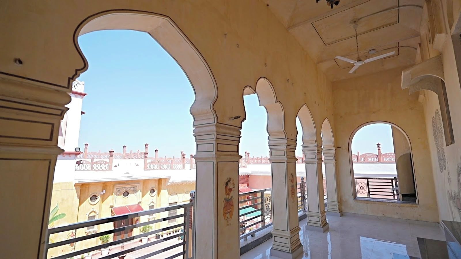 Corredor interno com arcos, ventilador de teto e vista para edifícios externos.