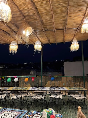 Uma área de refeições ao ar livre com mesas, cadeiras, um telhado de palha e luminárias pendentes.