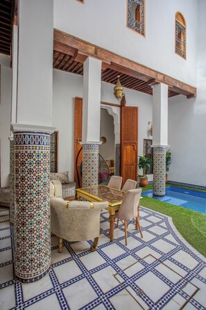 Uma área externa com mesas e cadeiras, cercada por colunas decoradas e um chão de azulejos.