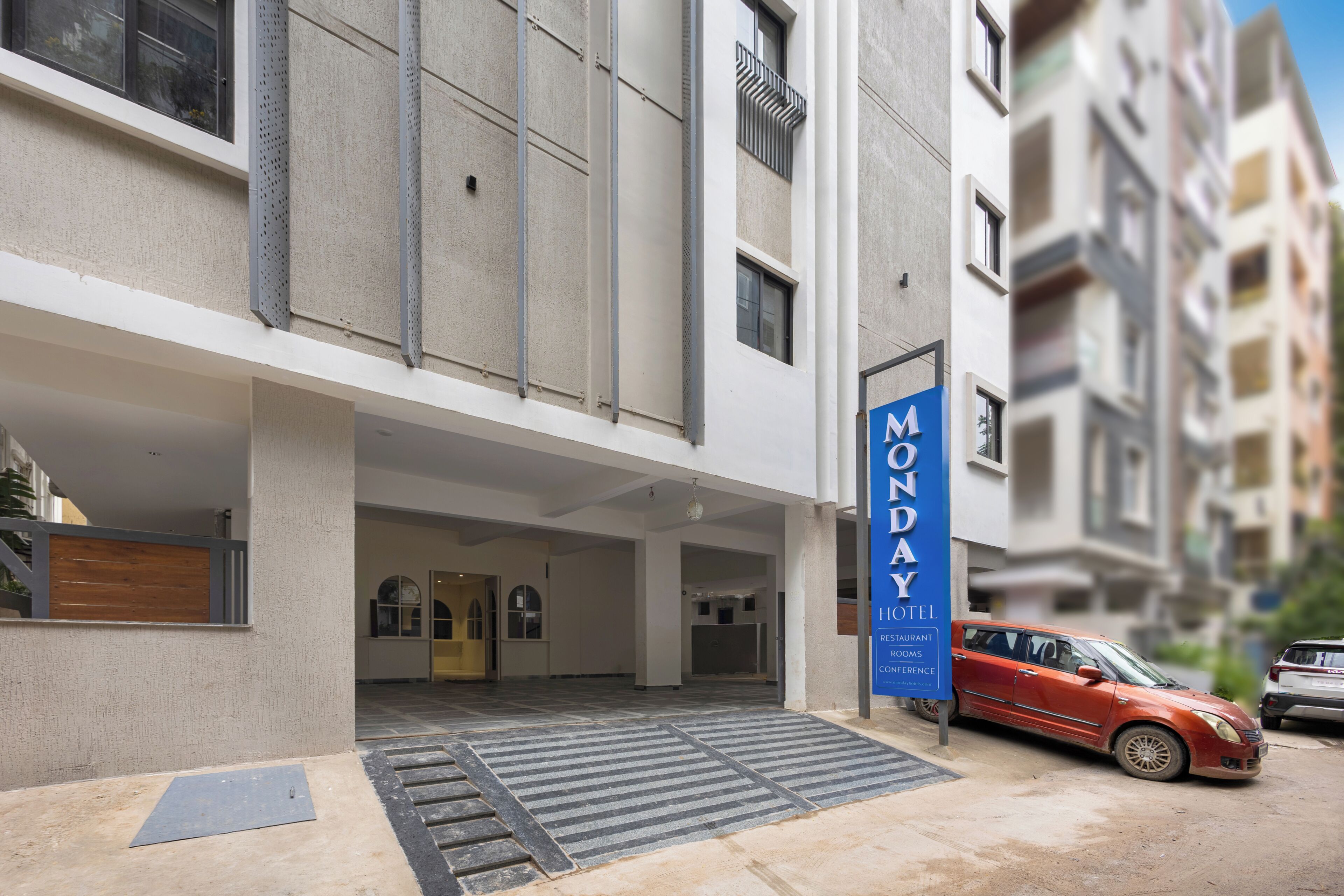 Entrada de um hotel com área externa coberta, escadas e um carro vermelho estacionado na frente.