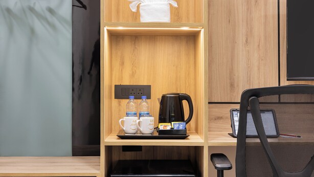 Quarto de hotel com prateleira de madeira contendo cafeteira, duas garrafas de água e duas canecas brancas com a inscrição 'hello Monday'.