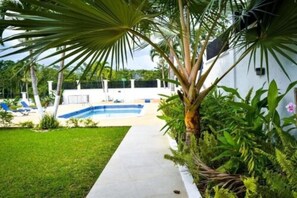 Uma área de piscina com um caminho acessível, rodeada por vegetação exuberante e um edifício branco.