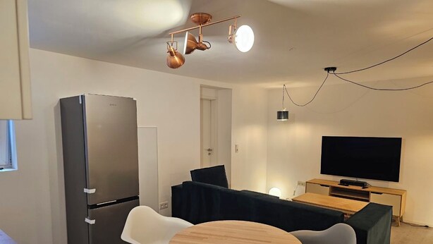 Um espaço compacto com cozinha compacta, área de refeições com mesa redonda e cadeiras, televisão de tela plana e luminária instalada no teto.