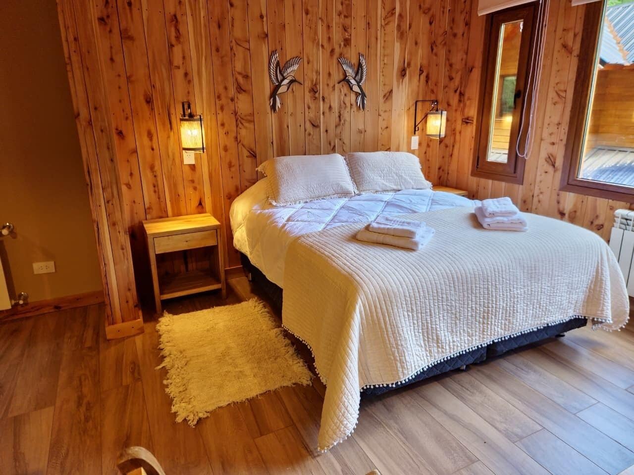 Quarto com painéis de madeira, uma cama, mesas de cabeceira e luzes fixadas na parede.