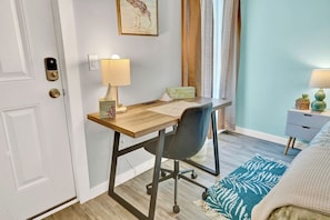Quarto com uma escrivaninha de madeira, uma cadeira, uma mesa de cabeceira com abajur, uma cama com manta de padrão azul e branco, e uma janela com cortinas.