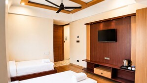Quarto de hotel com duas camas, ventilador de teto, televisão presa a um painel de madeira e cafeteira no aparador.