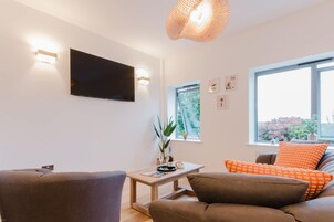 Uma sala de estar moderna com uma televisão de tela plana, uma mesa de centro de madeira e um sofá com almofadas laranja.