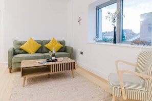 Uma sala de estar com um sofá verde, duas almofadas amarelas, uma mesa de centro de madeira, uma poltrona listrada e uma janela com vista para a cidade.