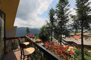Varanda com grade de madeira, duas cadeiras de madeira, mesa com frutas e bebidas, e vista para montanhas e casas.